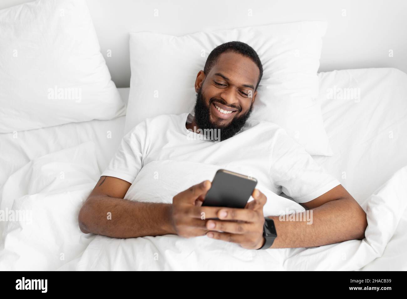Smiling young black bearded guy lies on white bed in bedroom, checks ...