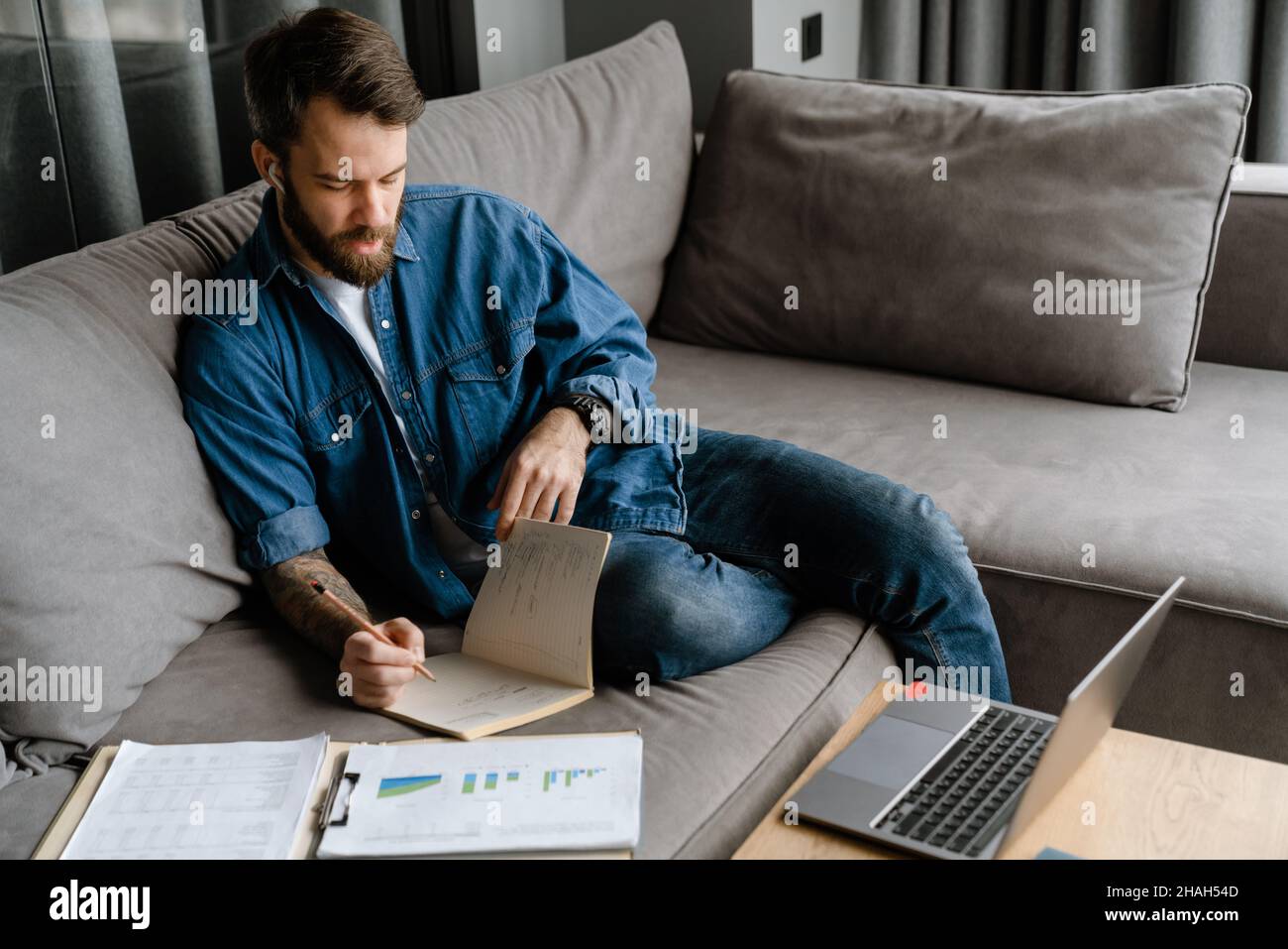 Bearded european man writing down notes while sitting on sofa at home ...