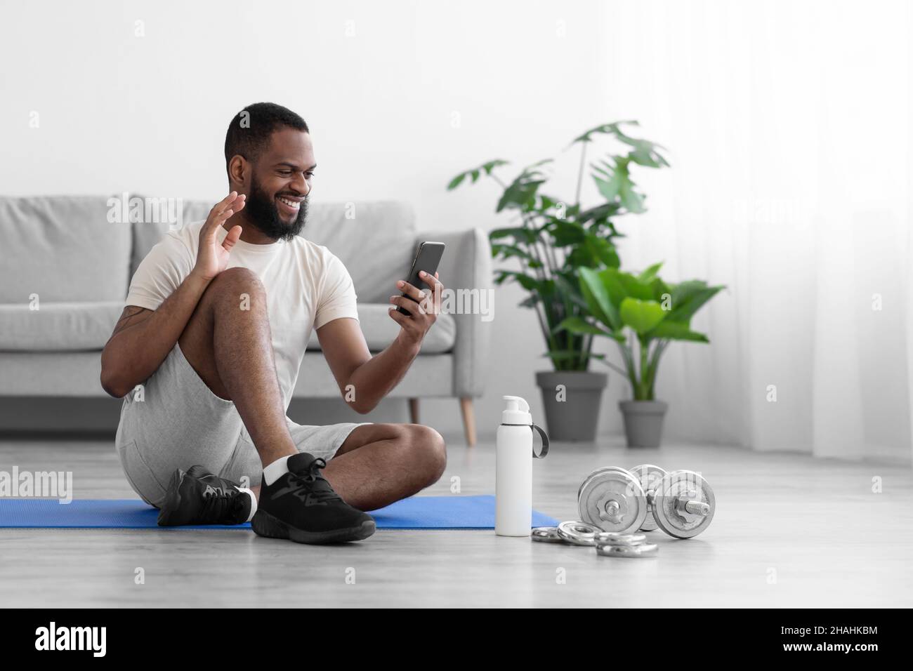 Cheerful young black guy sits on mat on floor in room interior with ...