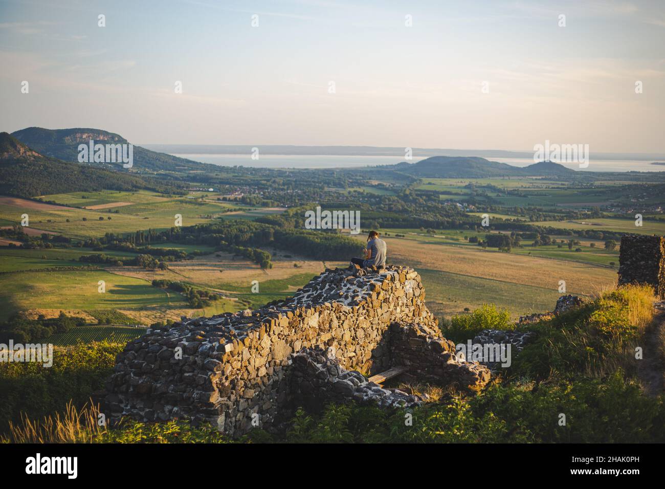 Female sitting on the half-ruined wall in the green field with a lake ...