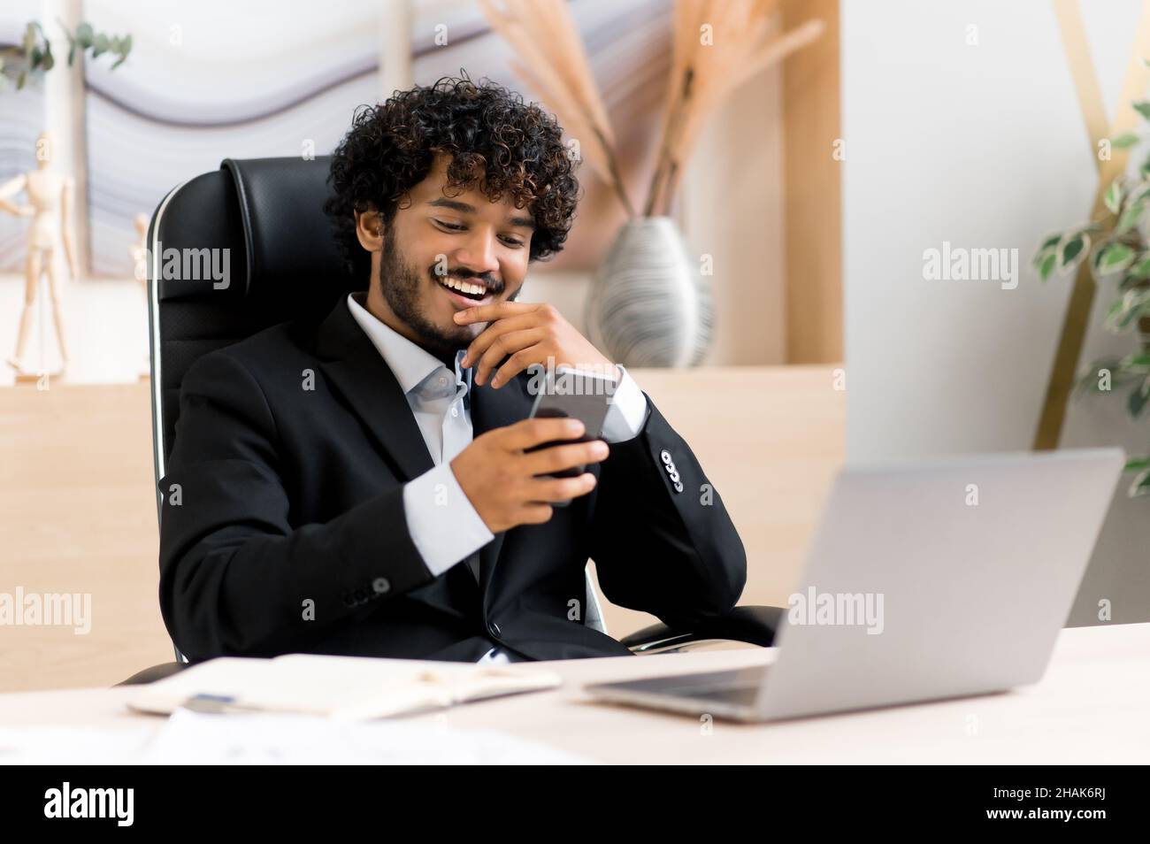 Successful joyful young Indian businessman, marketer or manager sitting at table in office ...