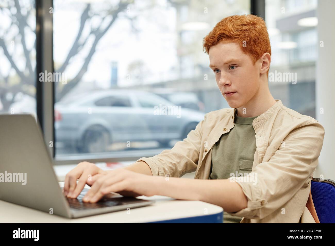 Portrait of red haired teenage boy using laptop in modern school ...