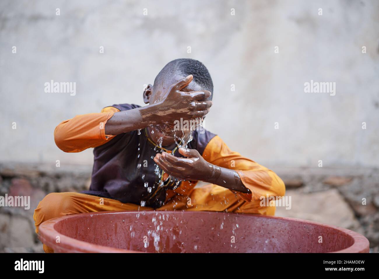 Close up one young black african boy wash face with fresh clean water from red bucket in hot day ...
