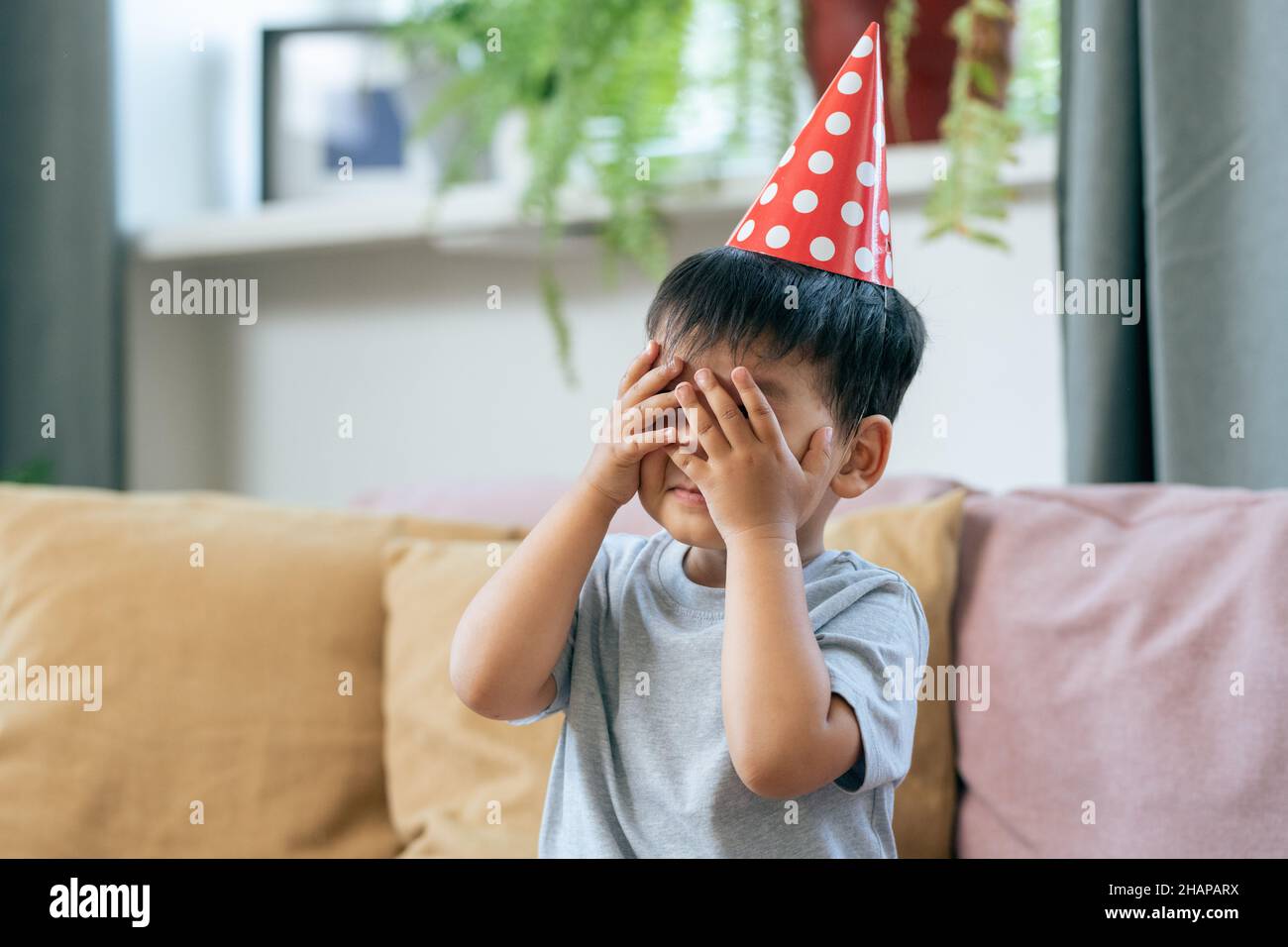 Cute little boy in birthday cap covering eyes with hands while sitting ...
