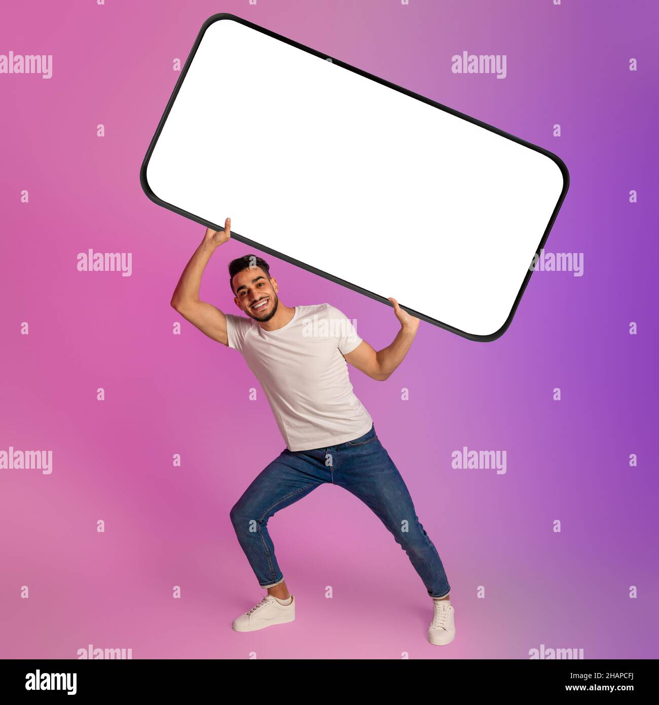 Cheerful young Arab man holding huge heavy smartphone with empty screen ...