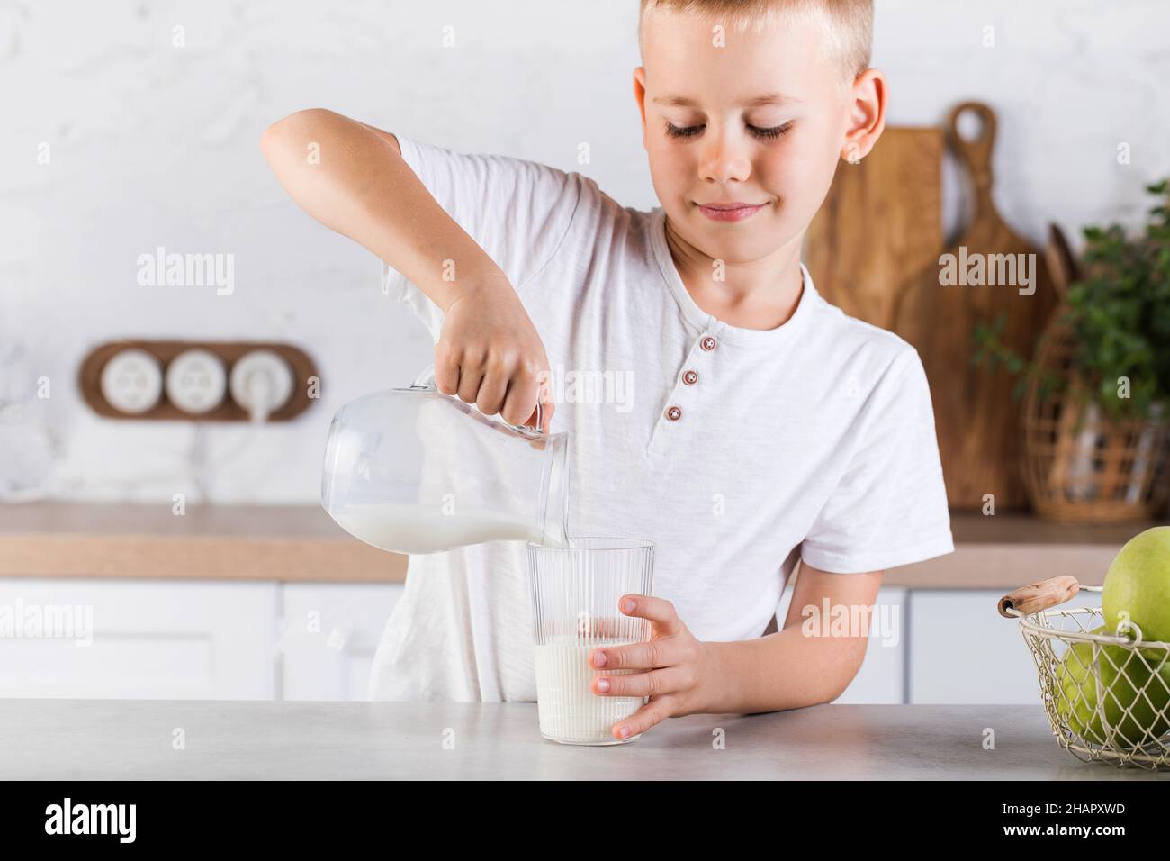 Cute smiling boy pours fresh milk from a jug into a mug in the kitchen ...