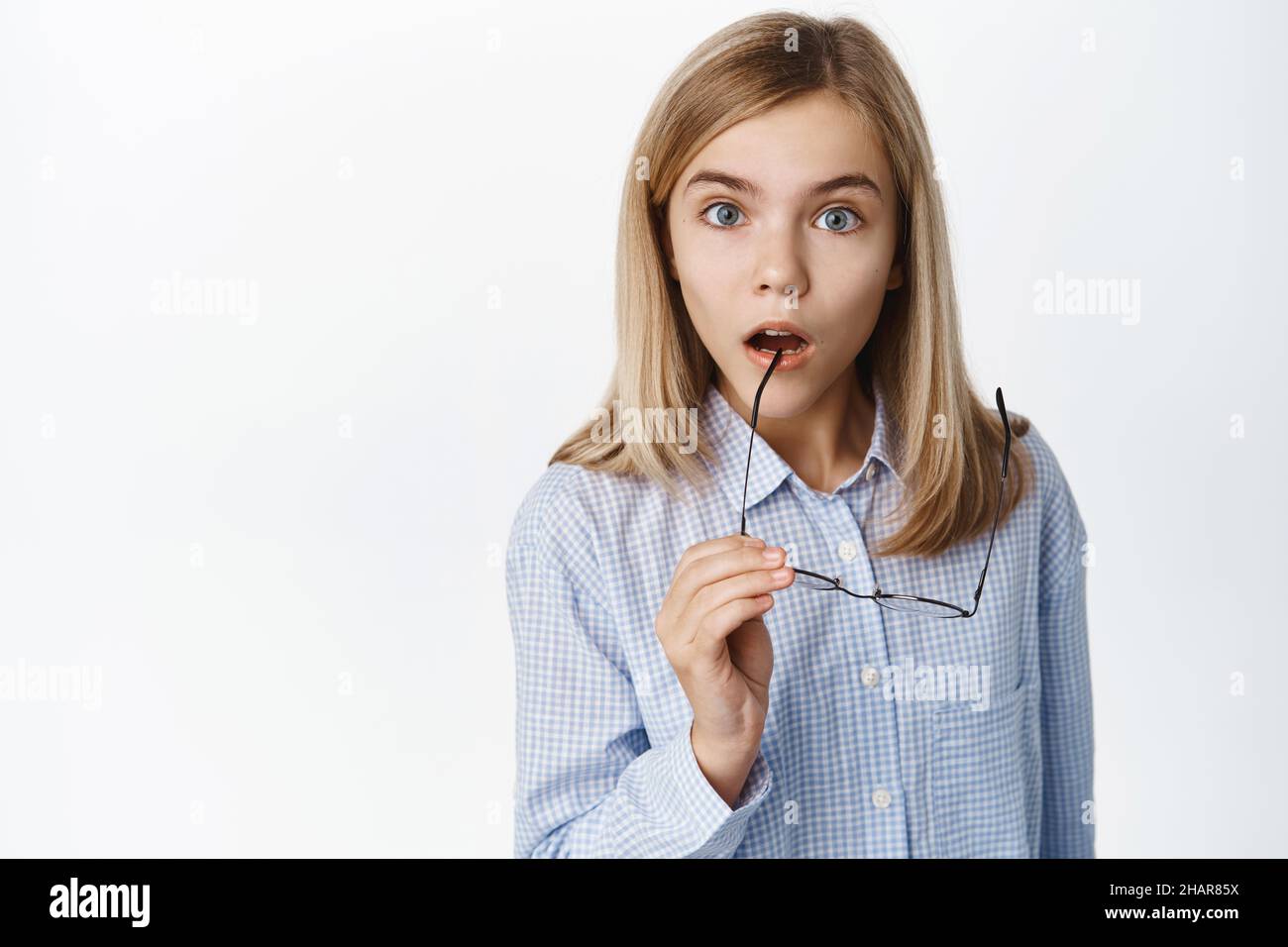 Portrait of cute blond girl, biting frame of glasses and looking amazed ...