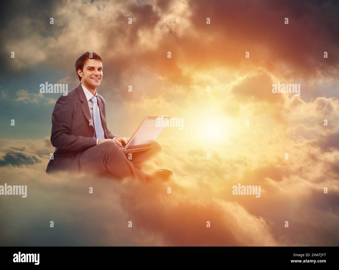 Happy businessman sitting on the clouds at sunset with his laptop ...