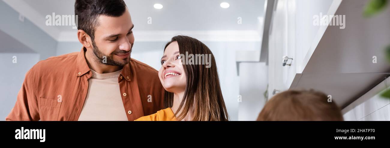 Smiling parents looking at each other near blurred kid in kitchen ...