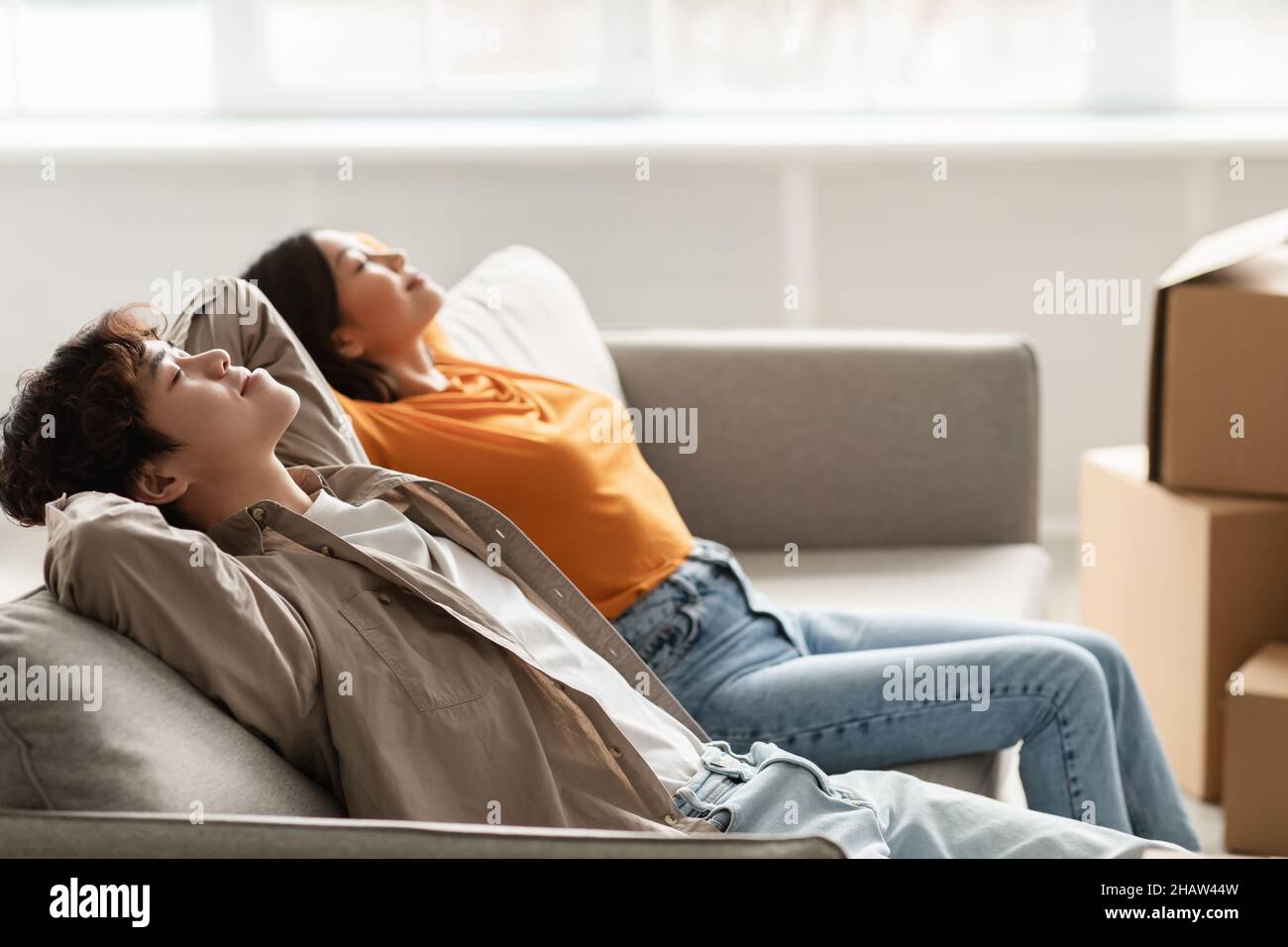 Positive young Asian family having rest on moving day, leaning back on ...