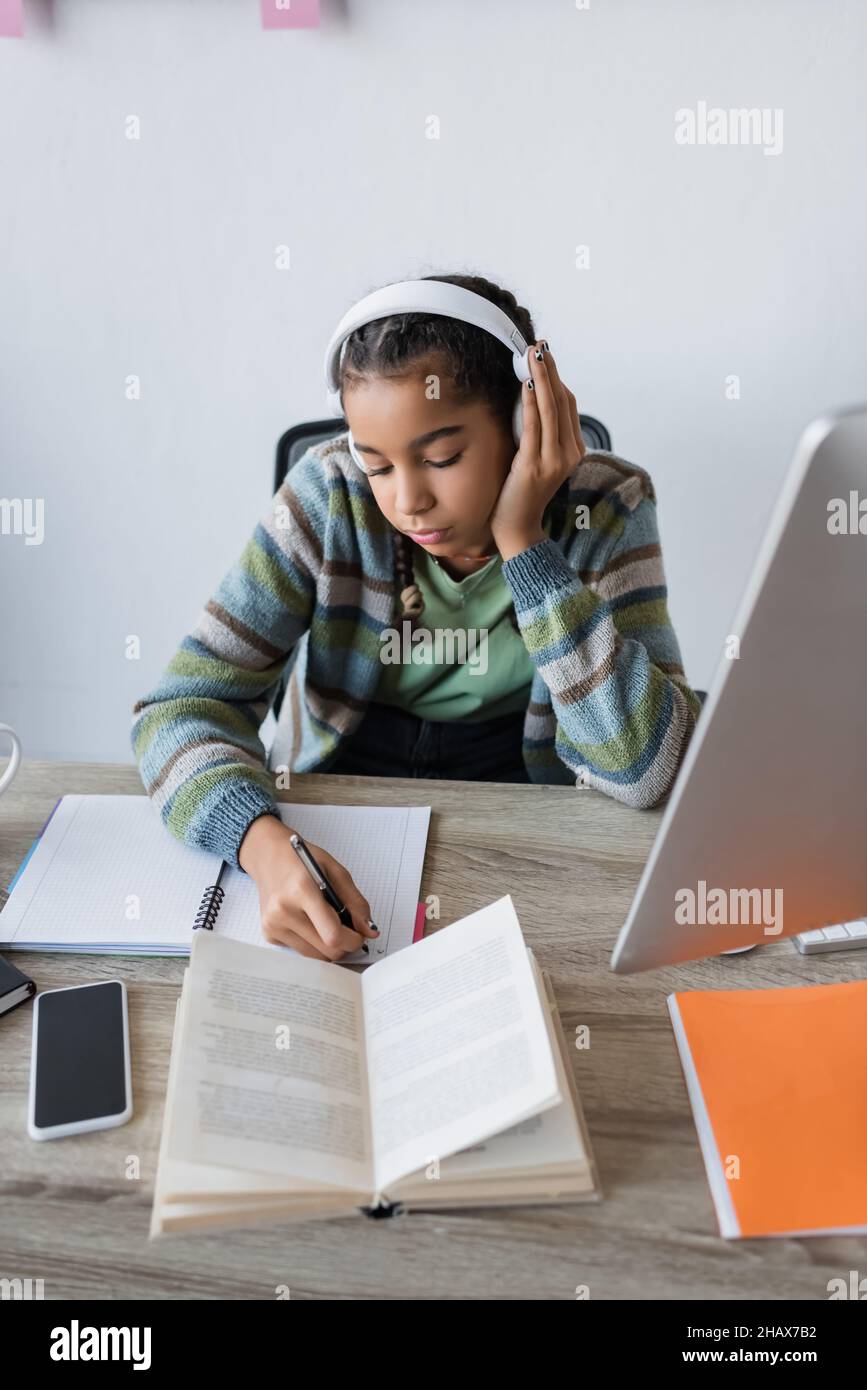african american girl in headphones writing in notebook near computer ...