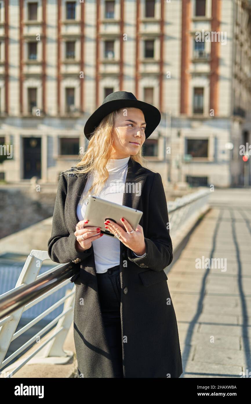 Young trendy female in black coat and hat surfing tablet while standing ...