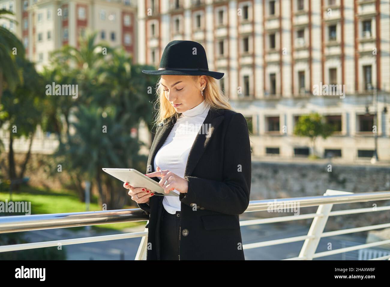 Side view of young trendy female in black coat and hat surfing tablet ...