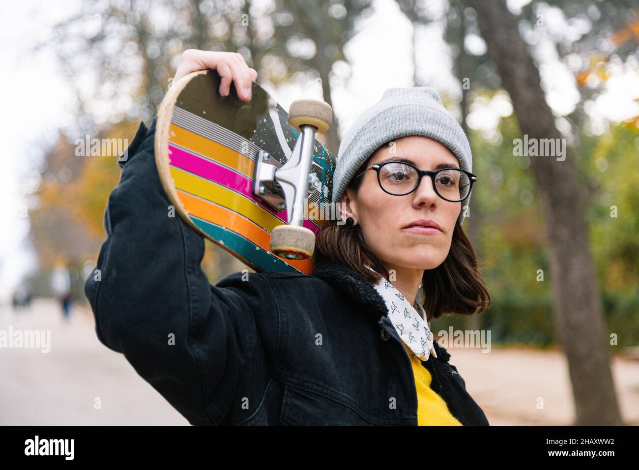Young female in black jacket with colorful skateboard behind neck ...