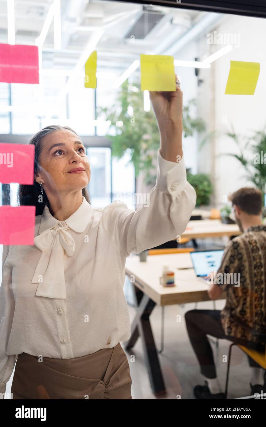 Happy female worker taking notes on stickers attached on glass wall of ...