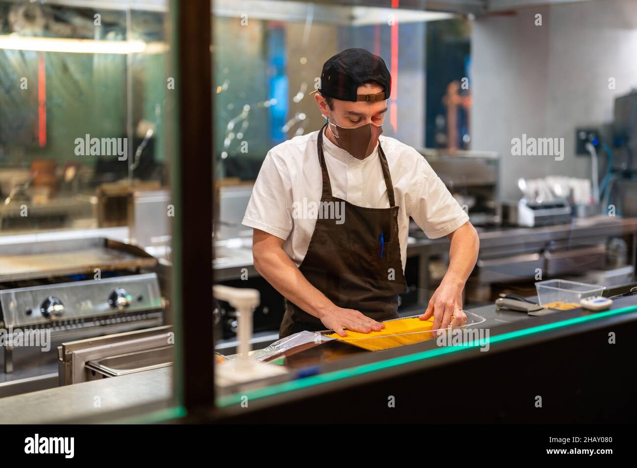 Side view of male cook in protective mask standing at stainless counter ...