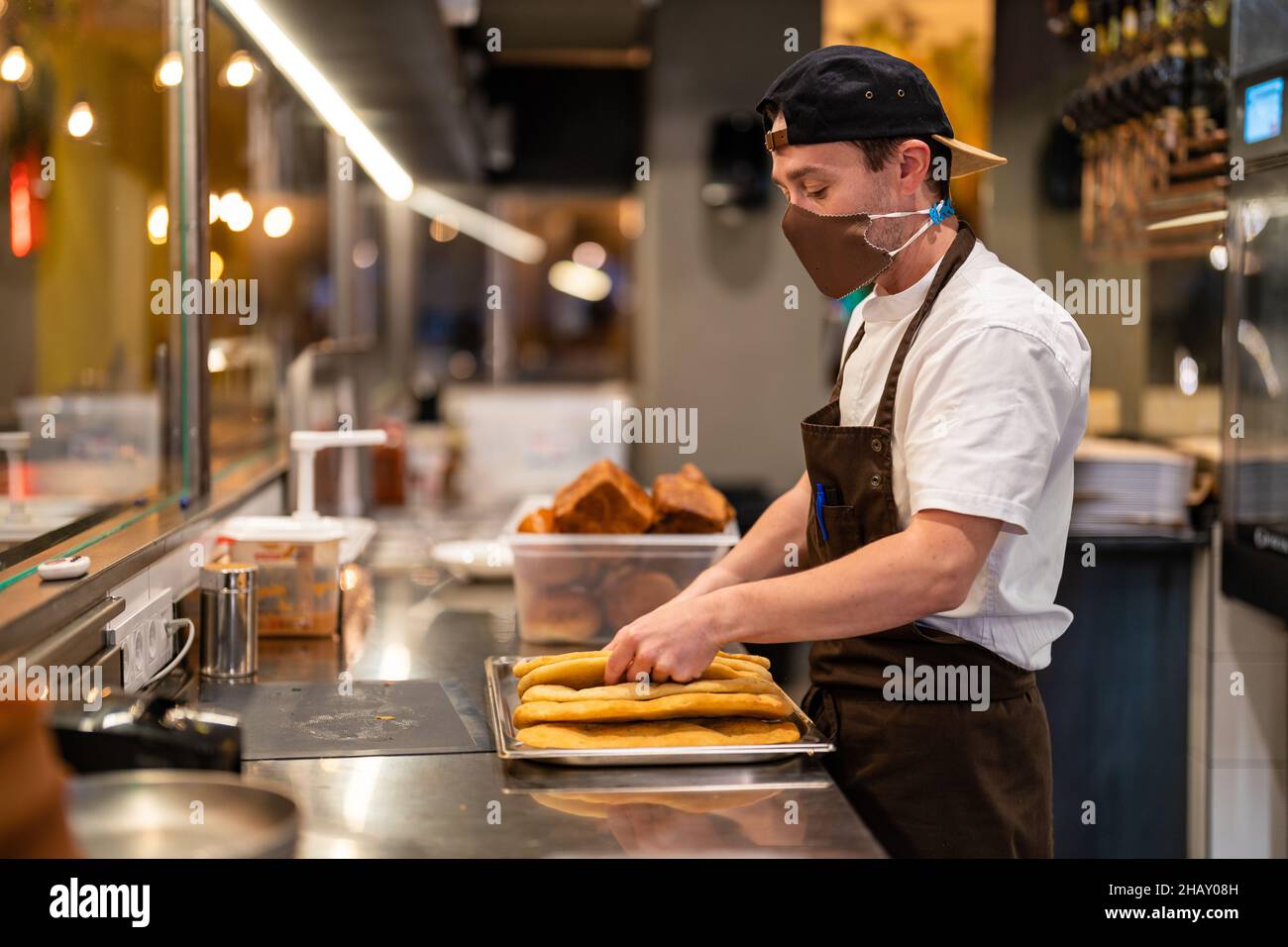Side view of male cook in protective mask standing at stainless counter ...