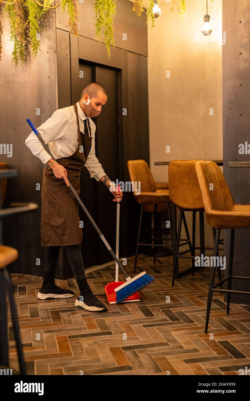 Full body of busy male waiter with scoop and broom cleaning floor in ...