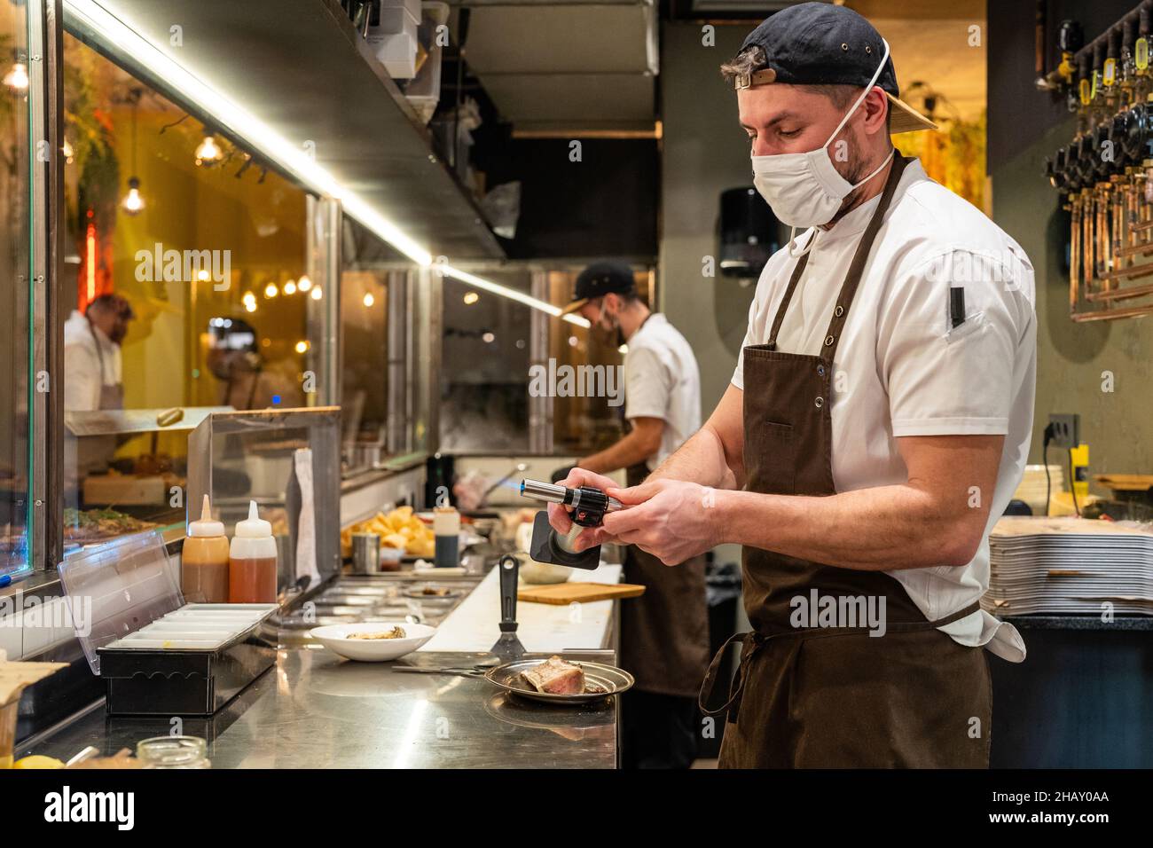 Side view of male cook in protective mask and apron using gas burner ...