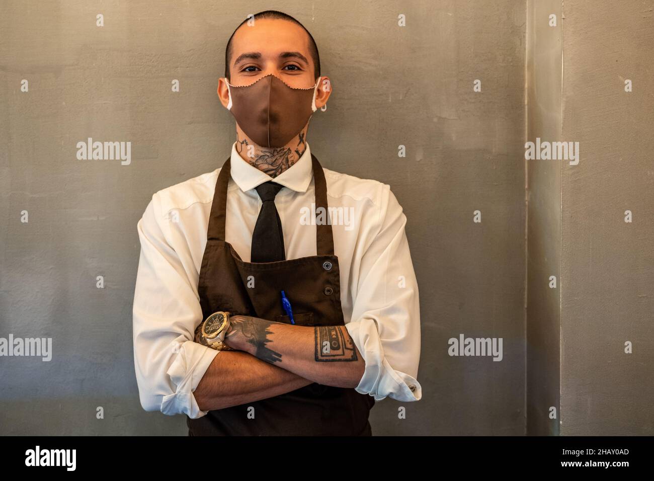 Male waiter with tattoos in white shirt and apron looking at camera ...
