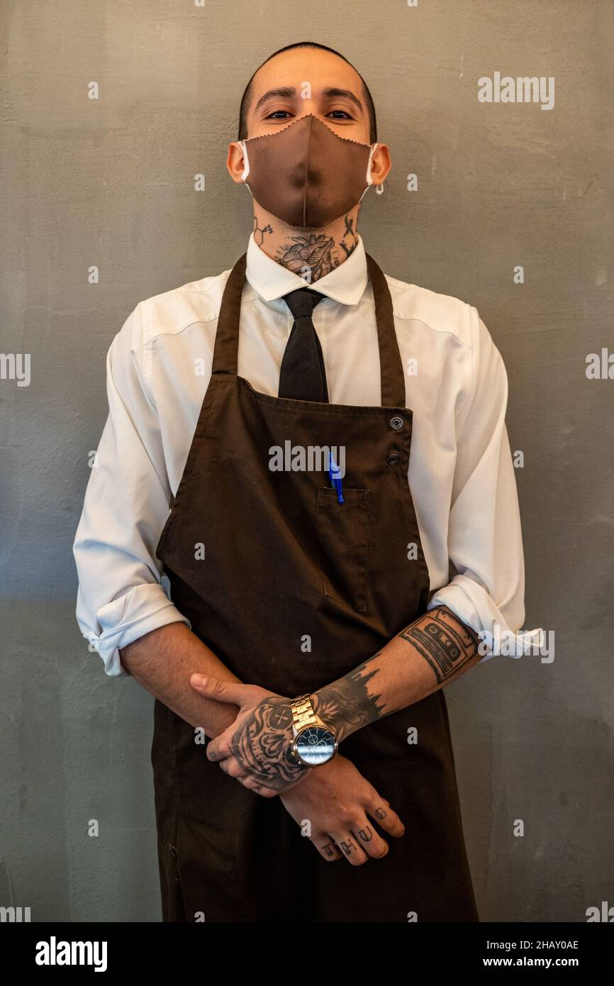 Male waiter with tattoos in white shirt and apron looking at camera ...