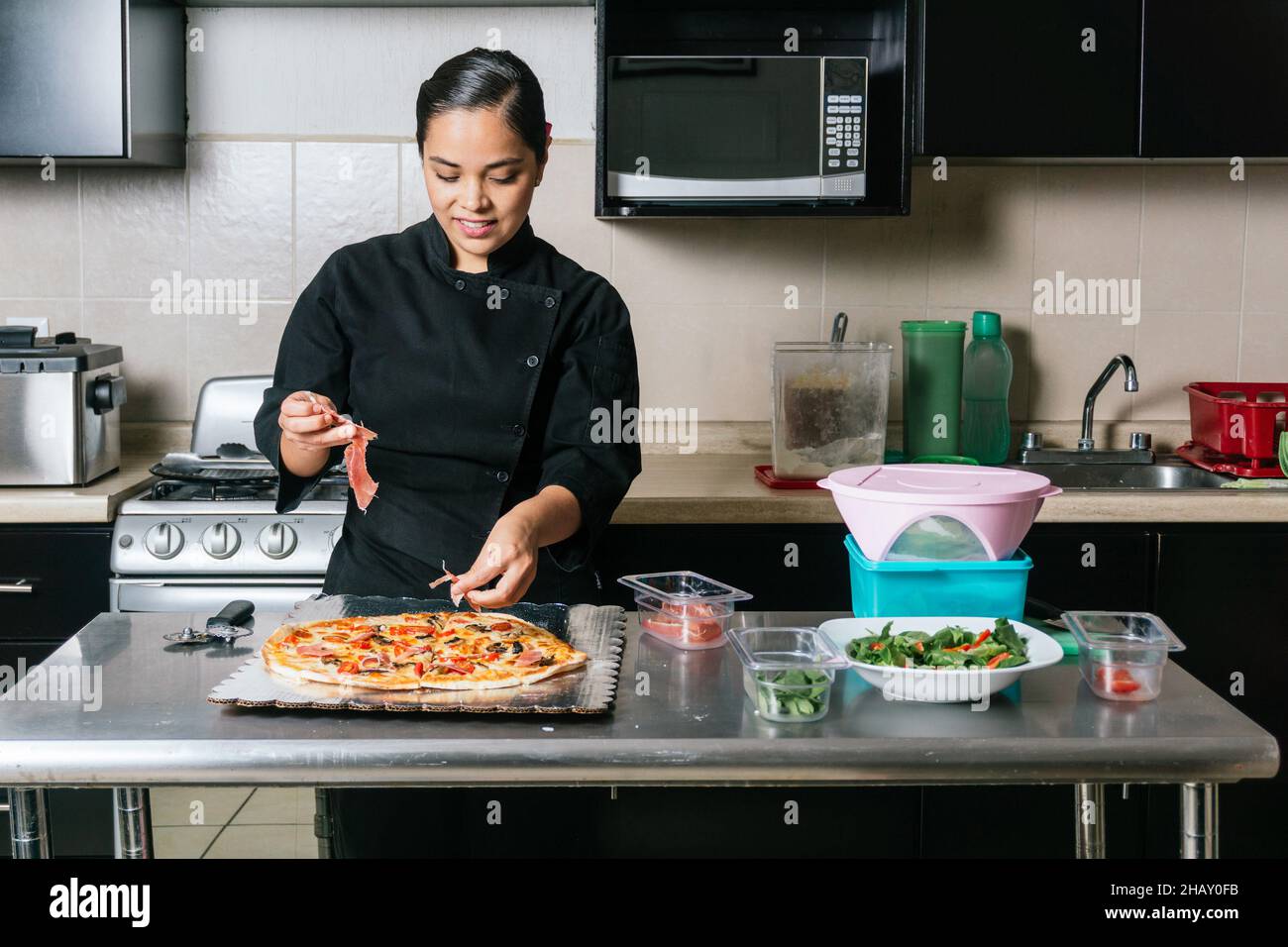Young ethnic female chef in uniform adding ham on delicious pizza while ...