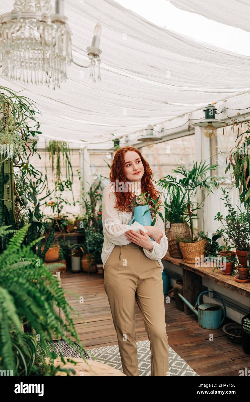 Charming female with long ginger hair standing with jug of seedling in ...