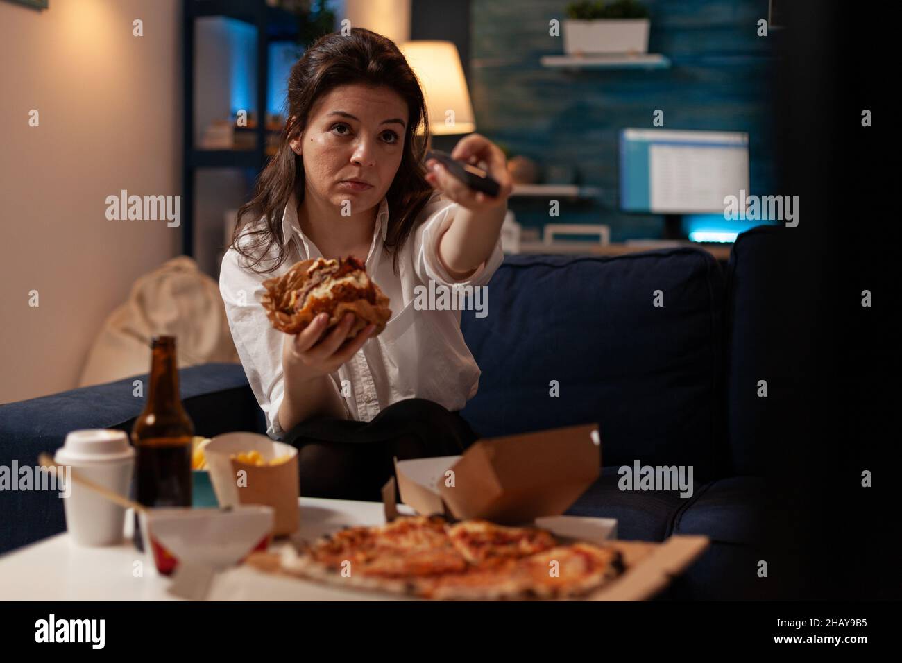 Young woman sitting on sofa eating double cheeseburger holding remote ...