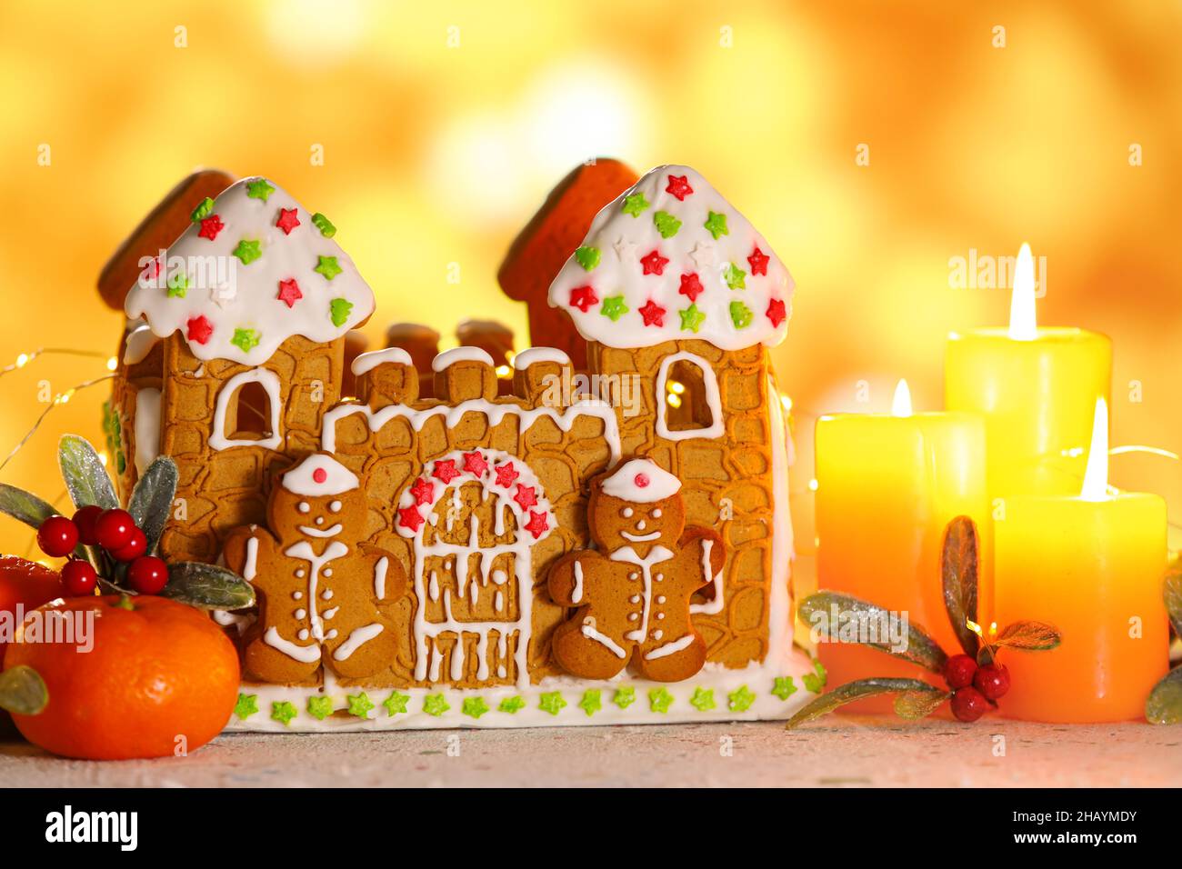 Gingerbread house, tangerine and burning candles on table against ...