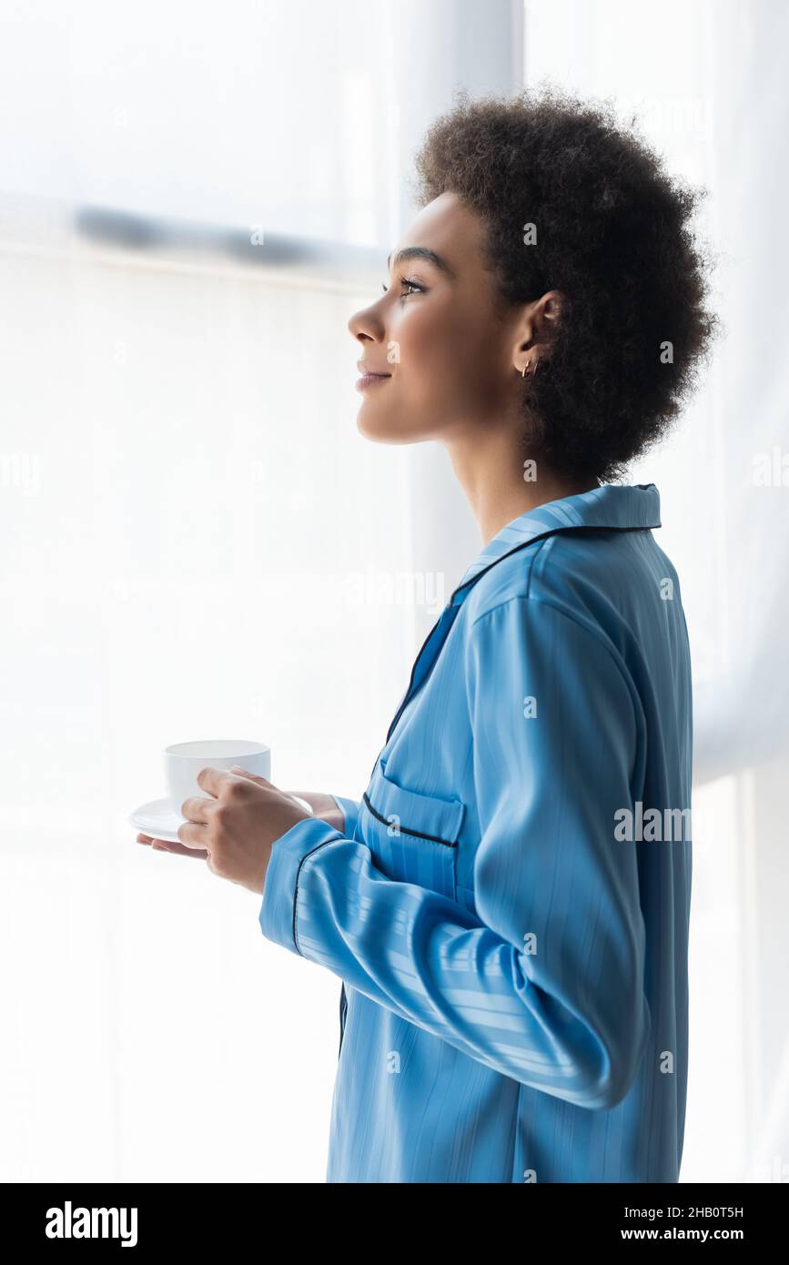 Side view of african american woman in pajamas holding cup near ...