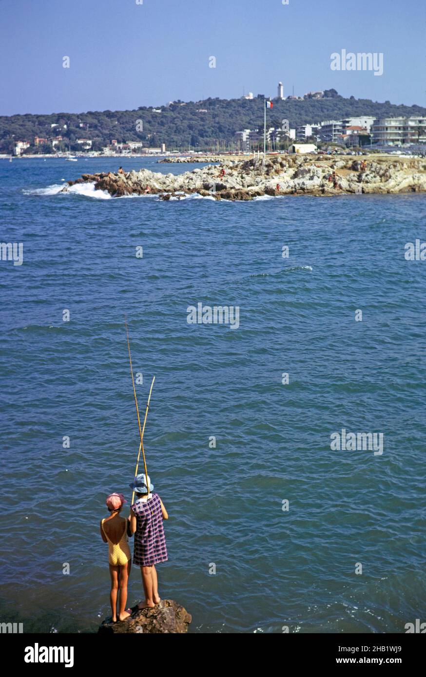 Two girls holding fishing rods standing on rock by the sea at Antibes ...