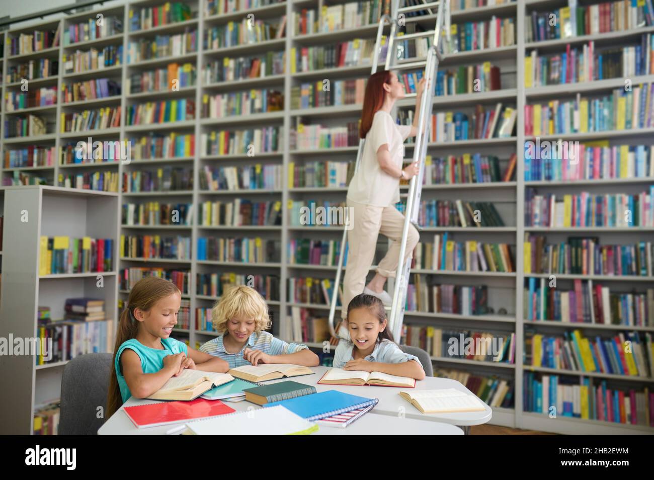 Children reading at table and woman on ladder near bookshelf Stock ...