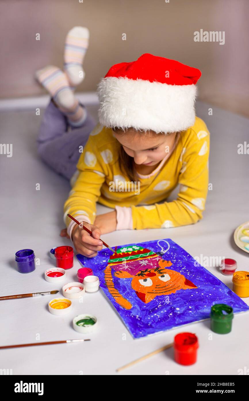 Little Girl in red santa claus hat lie on the floor and draw a ...