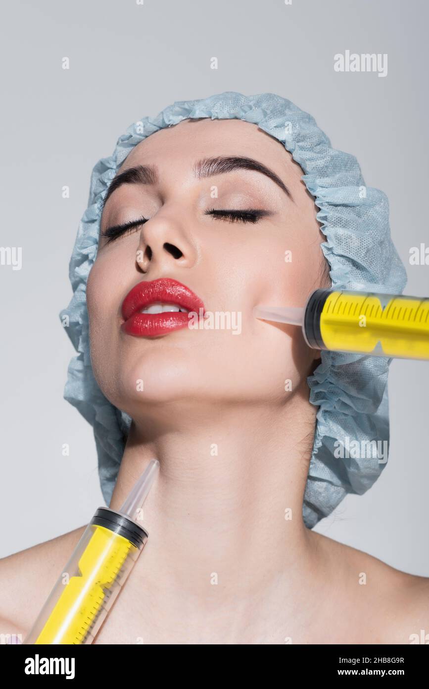 huge syringes near face of young woman in surgical cap isolated on grey ...