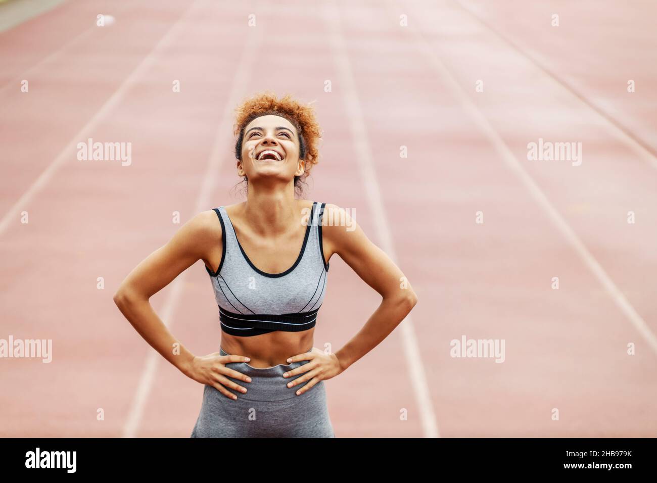 A happy sportswoman stands in the stadium with hands on hips and taking a break. Little by ...