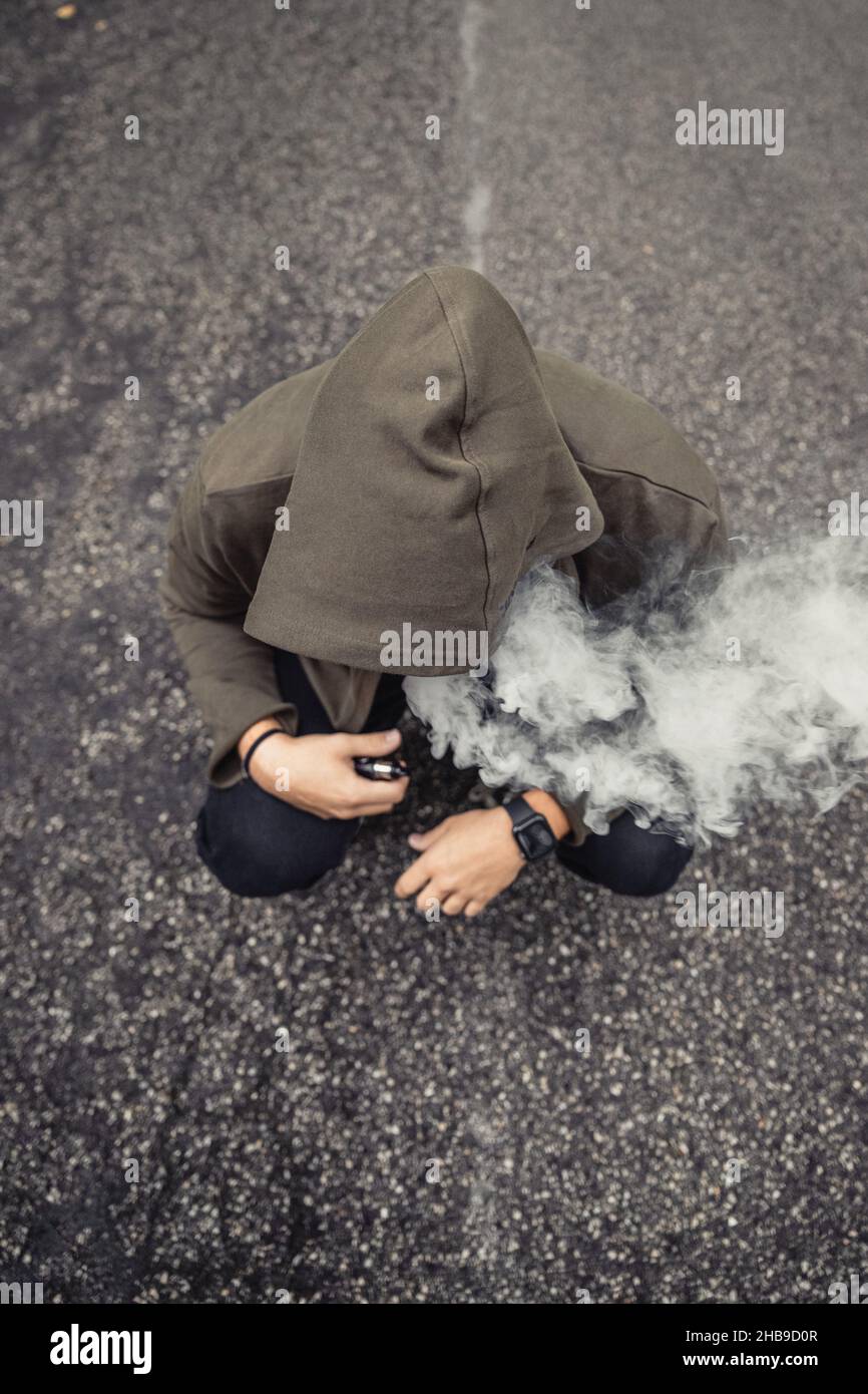 Vertical shot of the smoking male with the hood squatted outdoors in ...