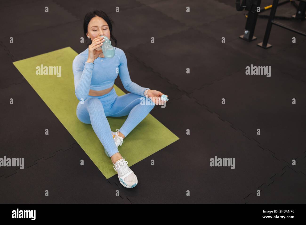 Sportive woman drinking water with bottle while sitting on mat at gym ...