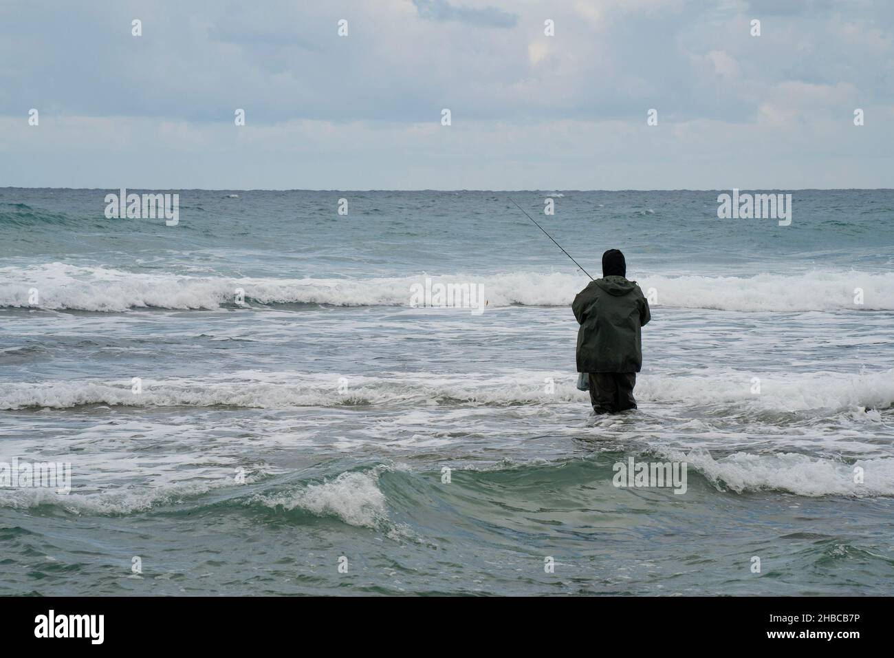 A lone fisherman standing knee deep in sea water on a stormy winter day ...