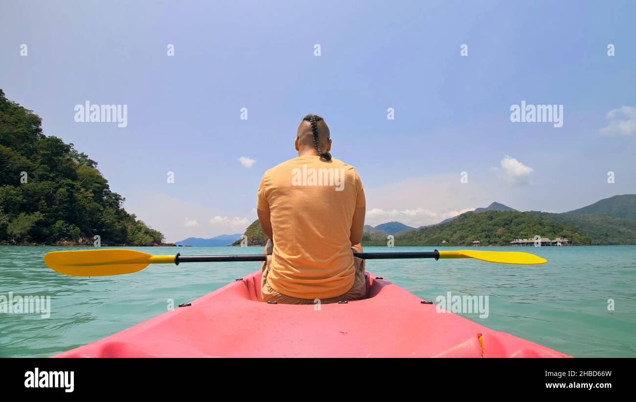 Young man with sunglasses and hat rows pink plastic canoe along sea ...