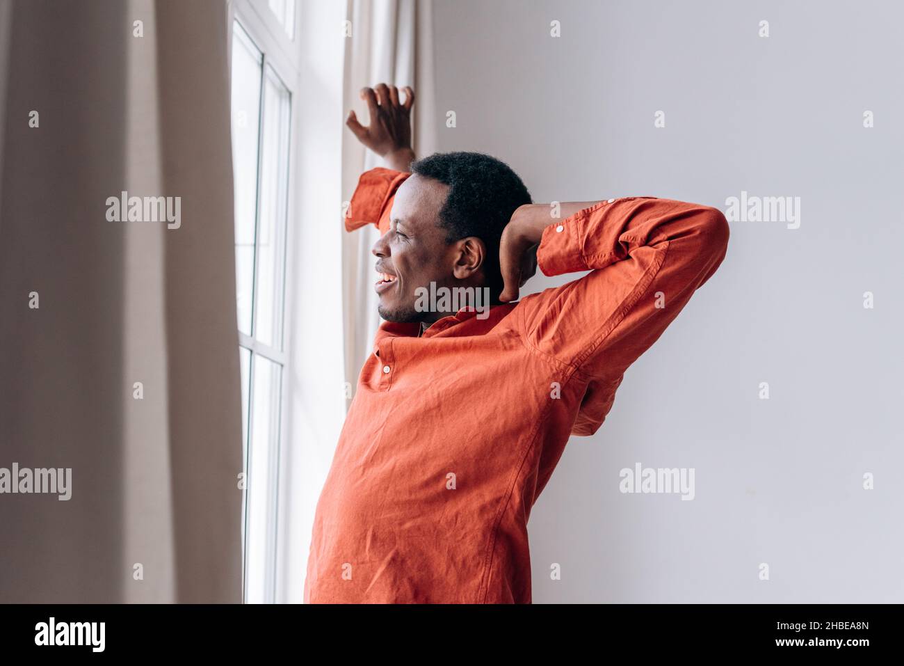 Joyful black young man in loose orange shirt stretches body looking out ...