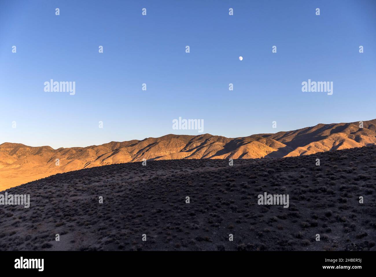 An early Moon shines over the mountains during the sunset in the desert ...