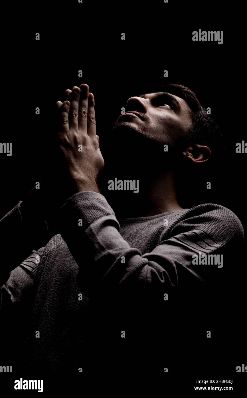 Headshot of serious confident young man praying in dark room. Lit from ...