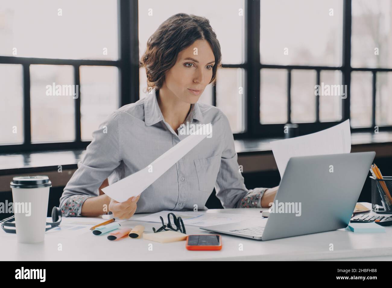 Focused hispanic woman worker analyzing documents and working on laptop ...