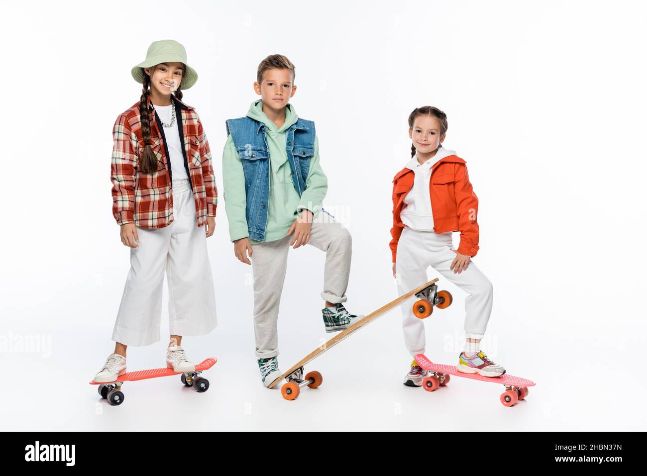 full length of trendy children smiling while standing with longboard ...
