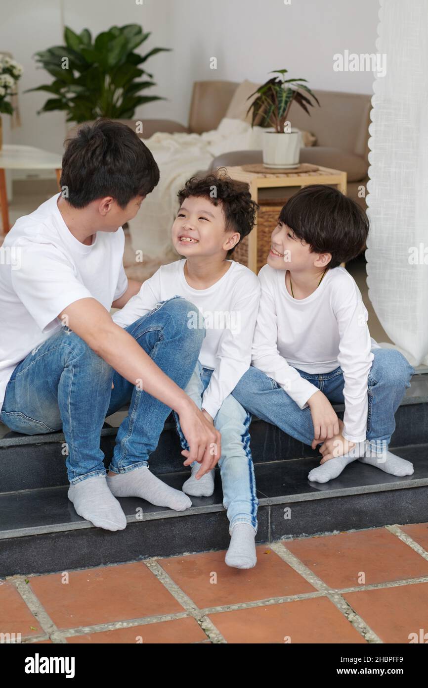 Joyful kids sitting on steps next to their joking father and looking at ...