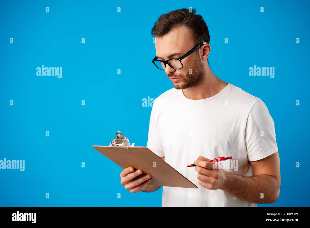 Portrait of young man making notes on clipboard against blue background ...