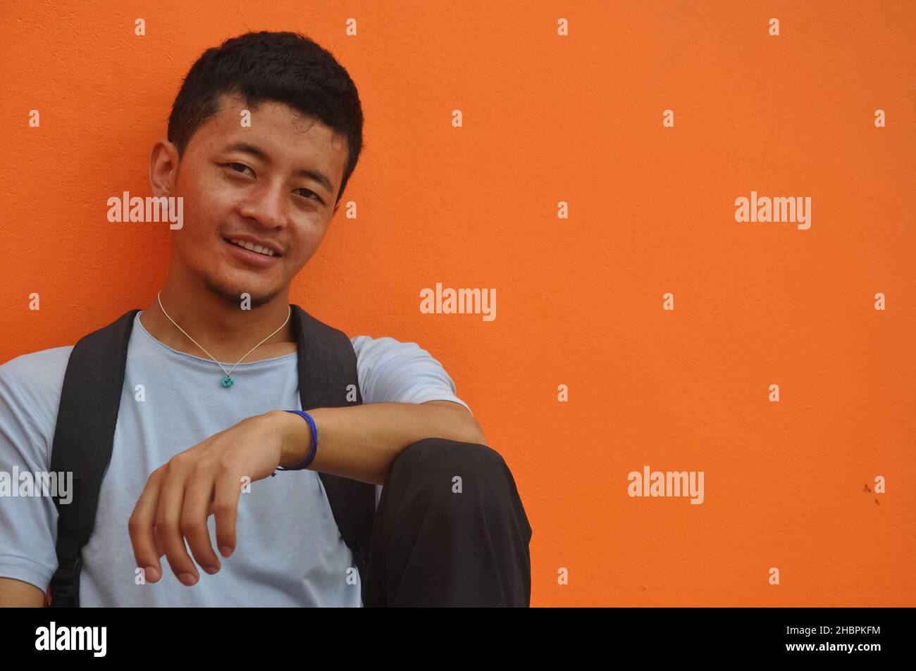 A happy Asian guy looking at camera, sitting against orange wall ...