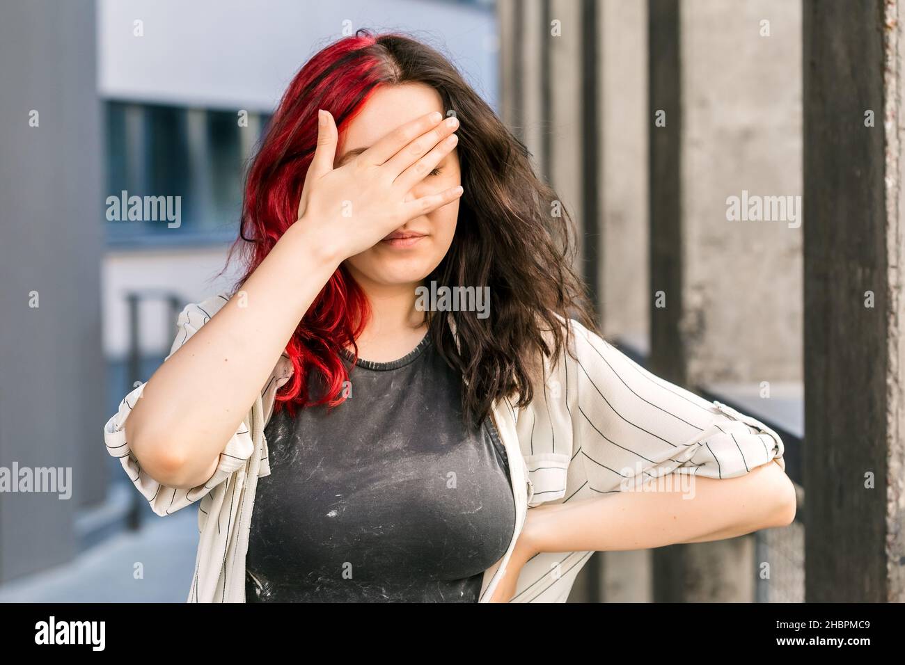 Stylish woman with red and black hair hiding eyes under hand while ...