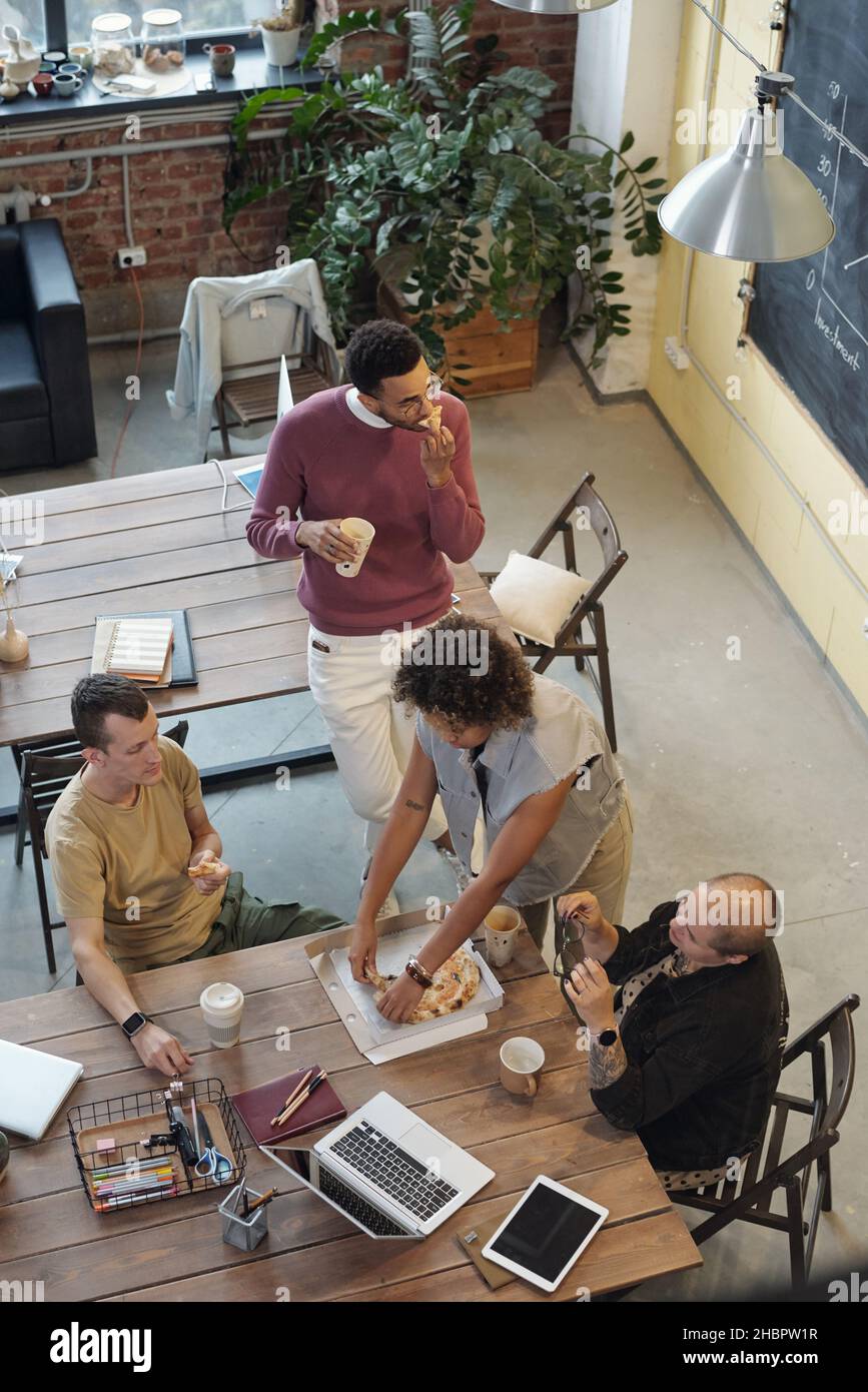 Four young co-workers gathered by table for lunch break eating pizza ...