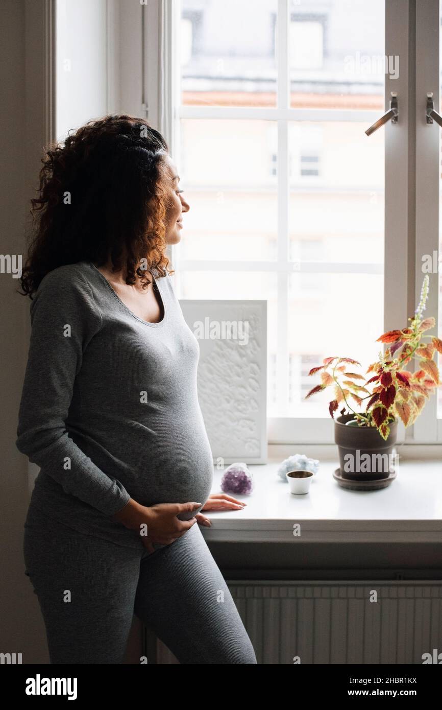 Pregnant woman with hand on stomach looking through window at home ...